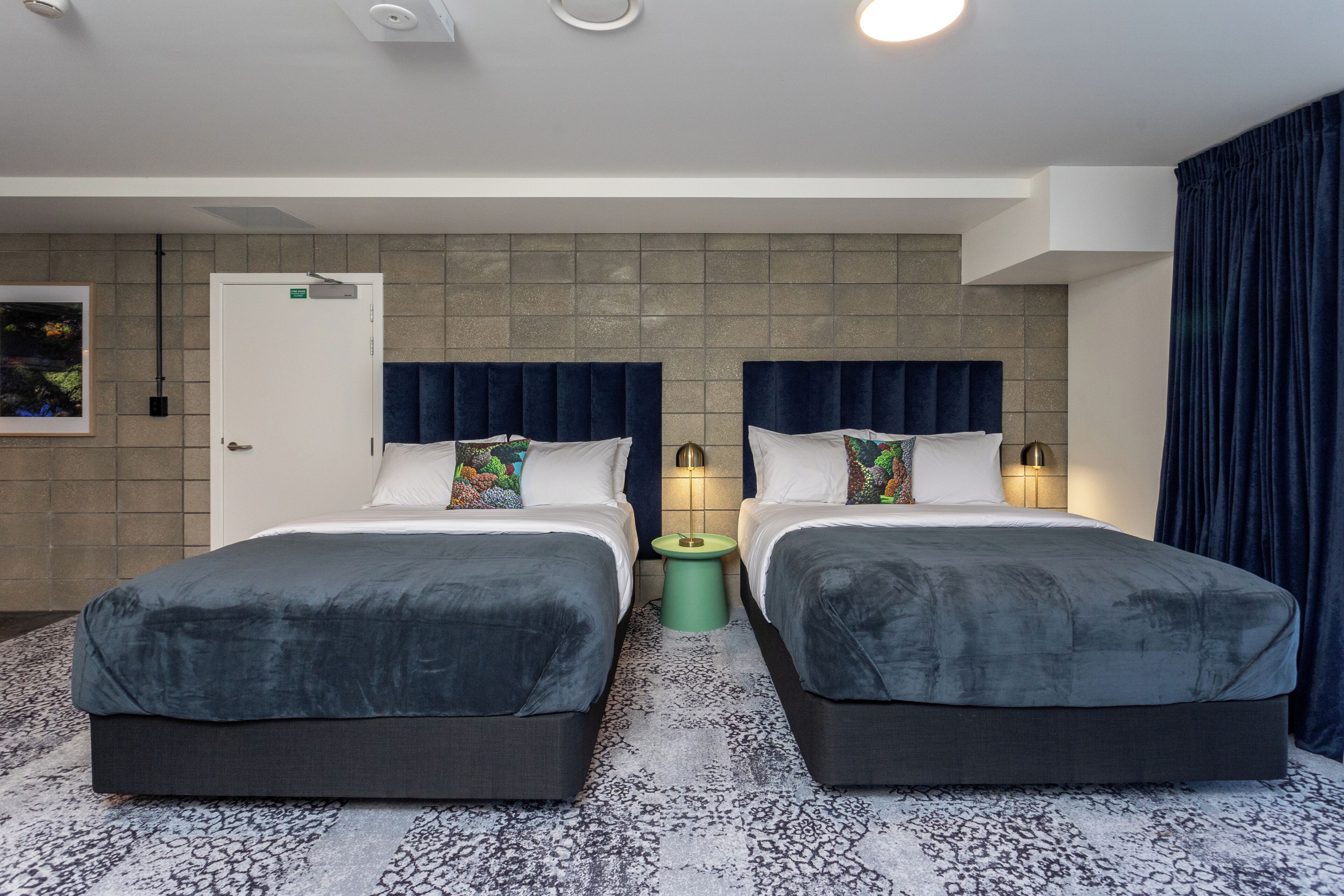 Quarto de hotel com duas camas, uma mesa de apoio verde e um tapete estampado.