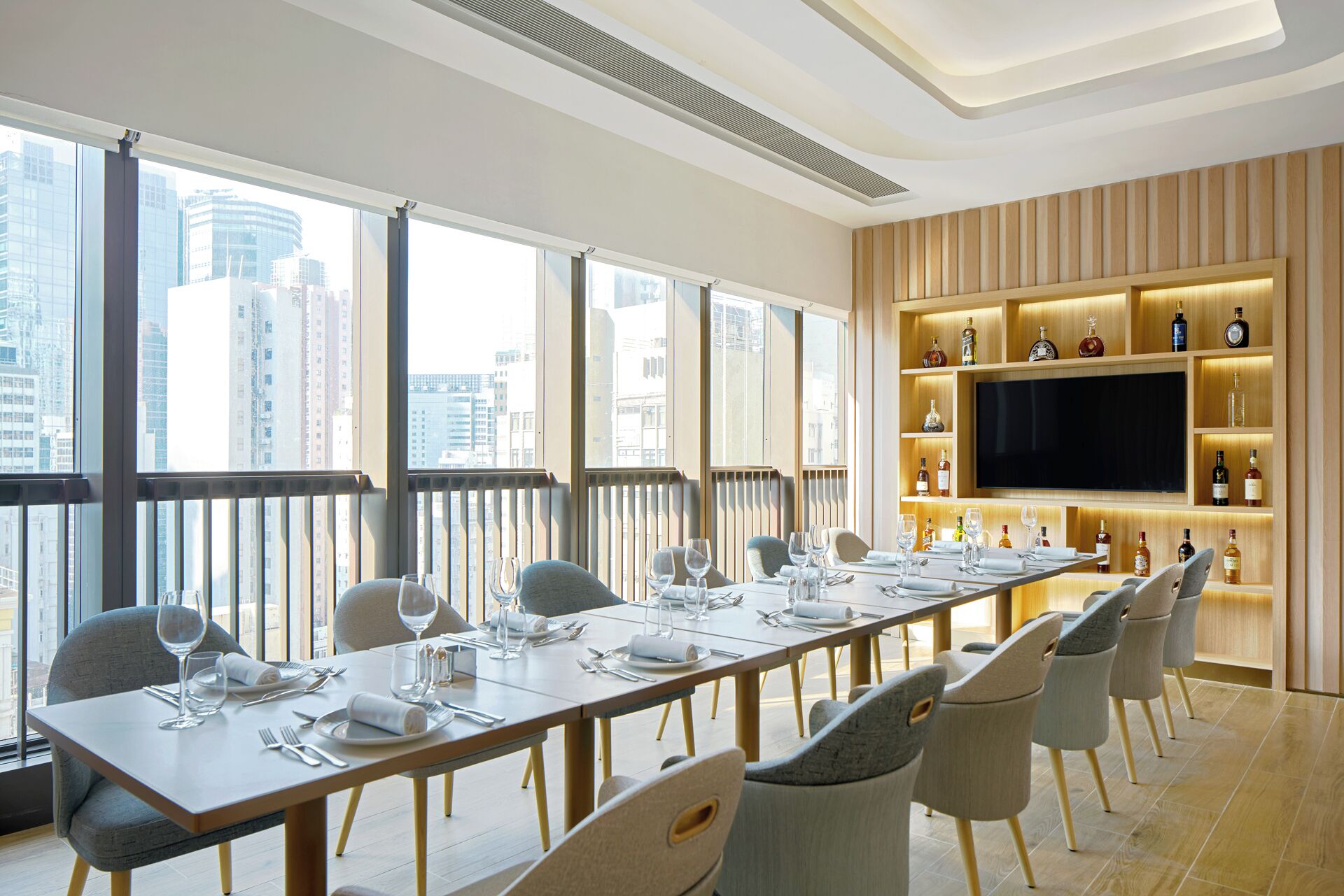 Uma área de jantar moderna com uma mesa longa posta para refeição, uma televisão na parede e vista da cidade através de grandes janelas.