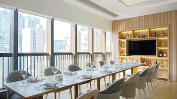 Uma área de jantar moderna com uma mesa longa posta para refeição, uma televisão na parede e vista da cidade através de grandes janelas.
