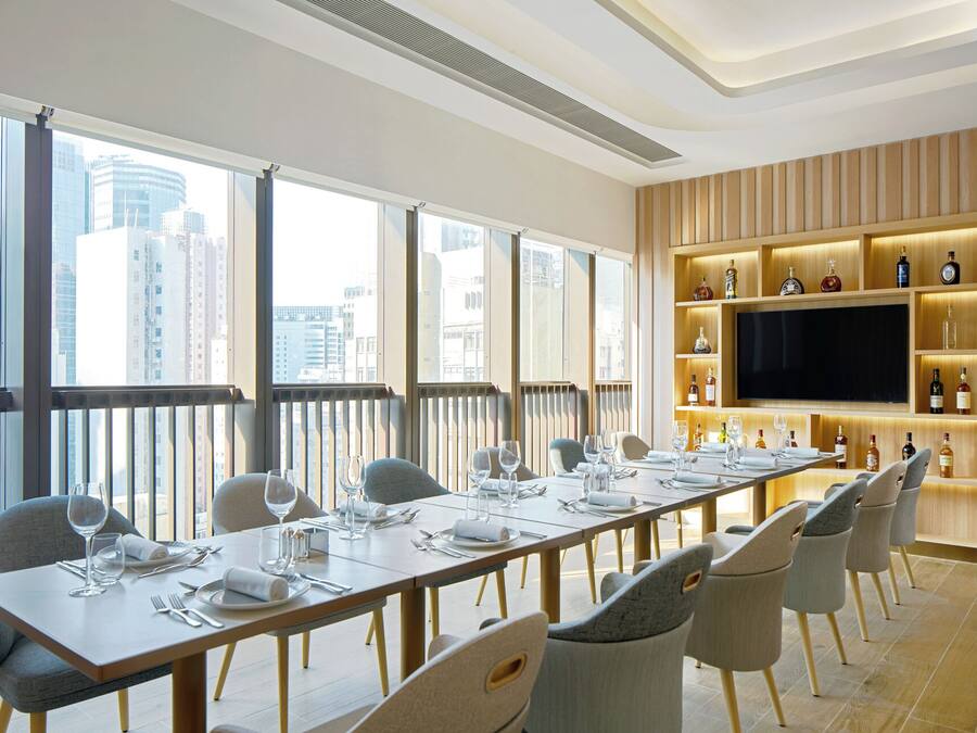 Uma área de jantar moderna com uma mesa longa posta para refeição, uma televisão na parede e vista da cidade através de grandes janelas.