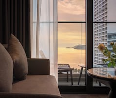 Uma sala de estar moderna com um sofá, uma mesa pequena e vista para o mar através de janelas amplas.
