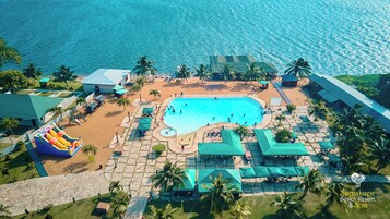 Vista aérea de um resort à beira-mar, com uma piscina grande, palmeiras e guarda-sóis.