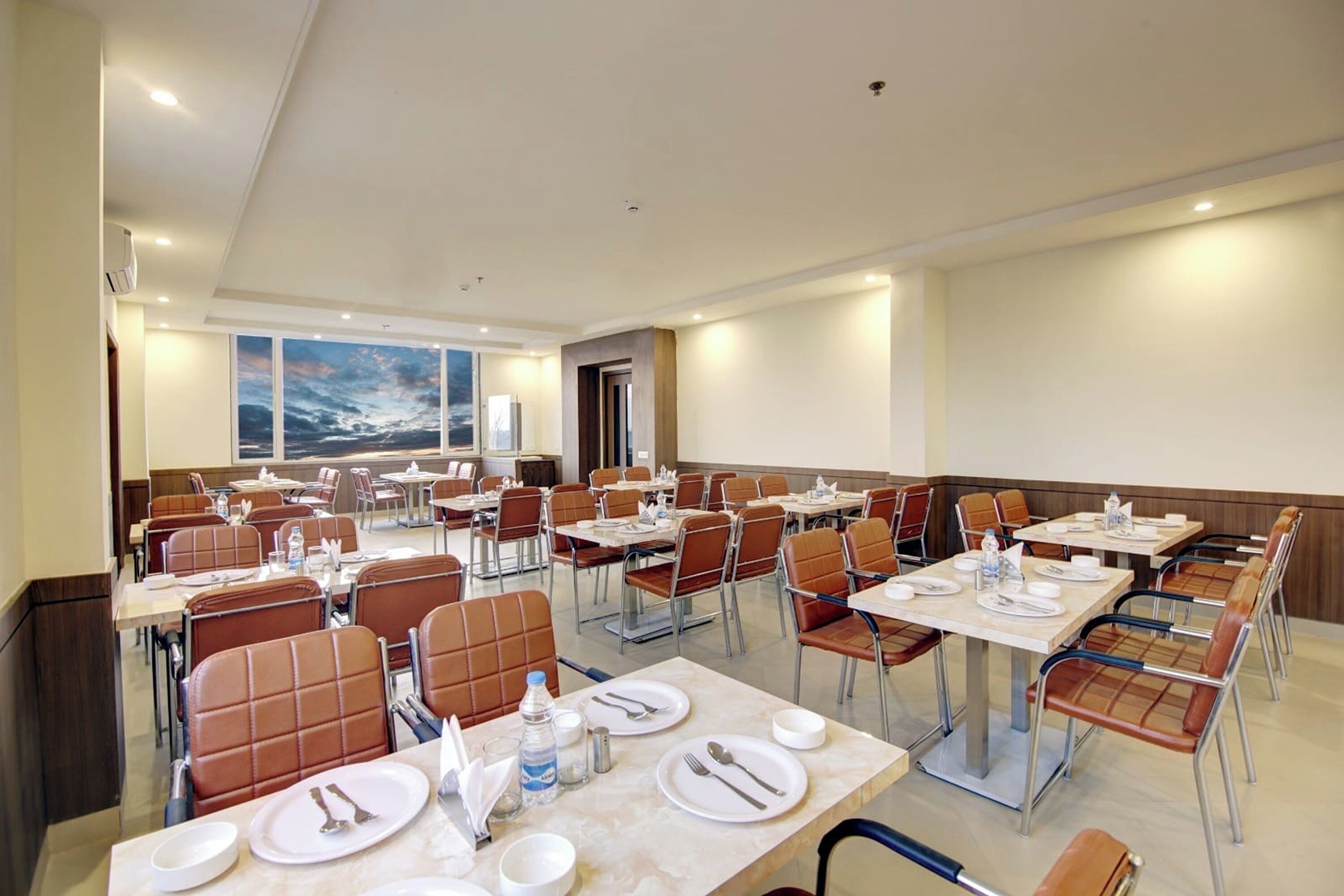 Uma sala de jantar de hotel com várias mesas e cadeiras, janelas amplas com vista e design de interiores moderno.