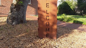 Uma placa de madeira com a palavra 'WELCOME' entalhada, colocada em frente a um edifício de tijolos e uma árvore próxima.