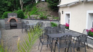 Um pátio externo com chão de tijolos, mesas e cadeiras de metal, churrasqueira de alvenaria e plantas em vasos.