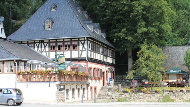 Um edifício tradicional em enxaimel com um telhado empena pronunciado, uma escadaria de pedra que leva à entrada e uma jardineira com flores vermelhas.