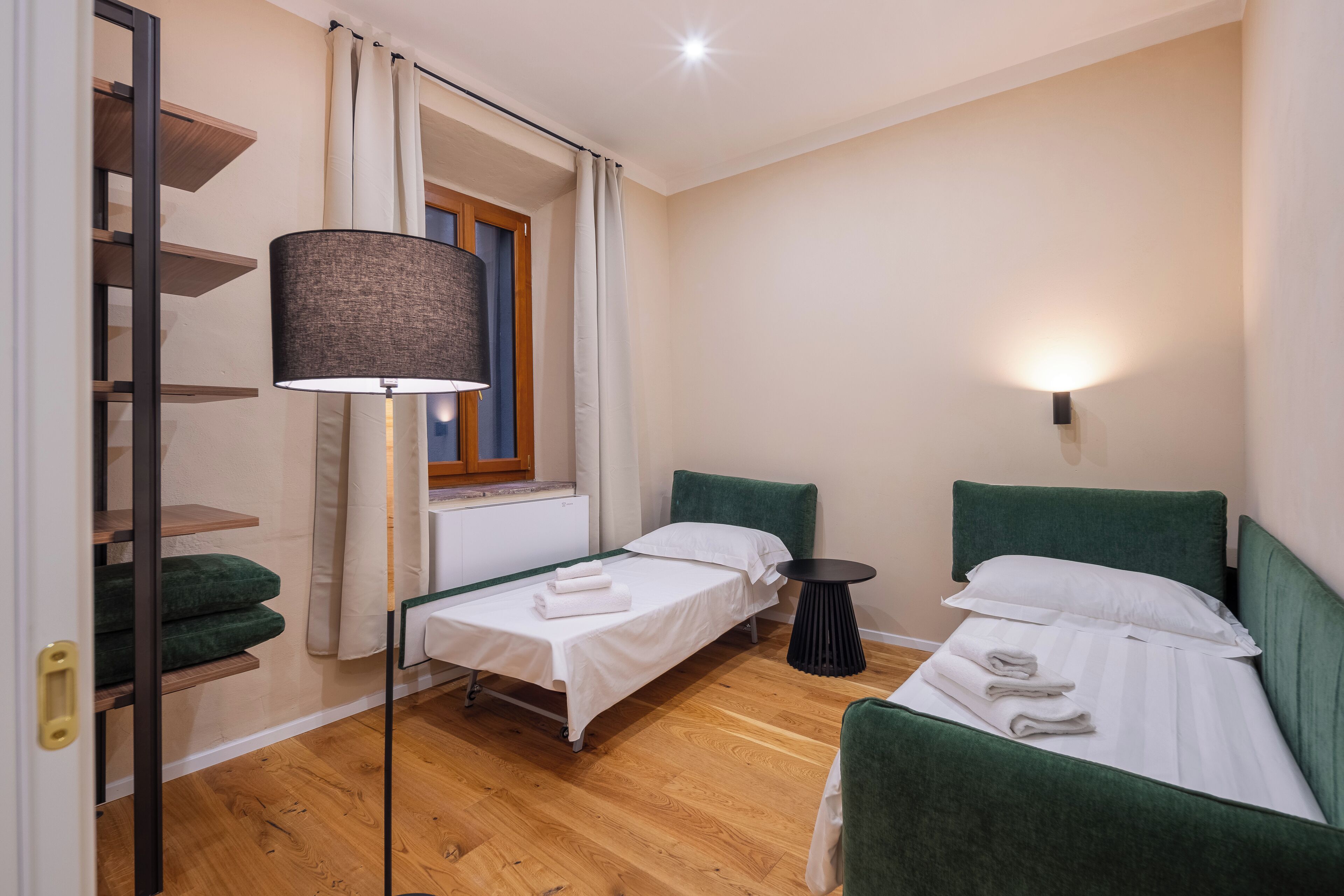 Um quarto pequeno e aconchegante com duas camas, um abajur e uma janela com cortinas.
