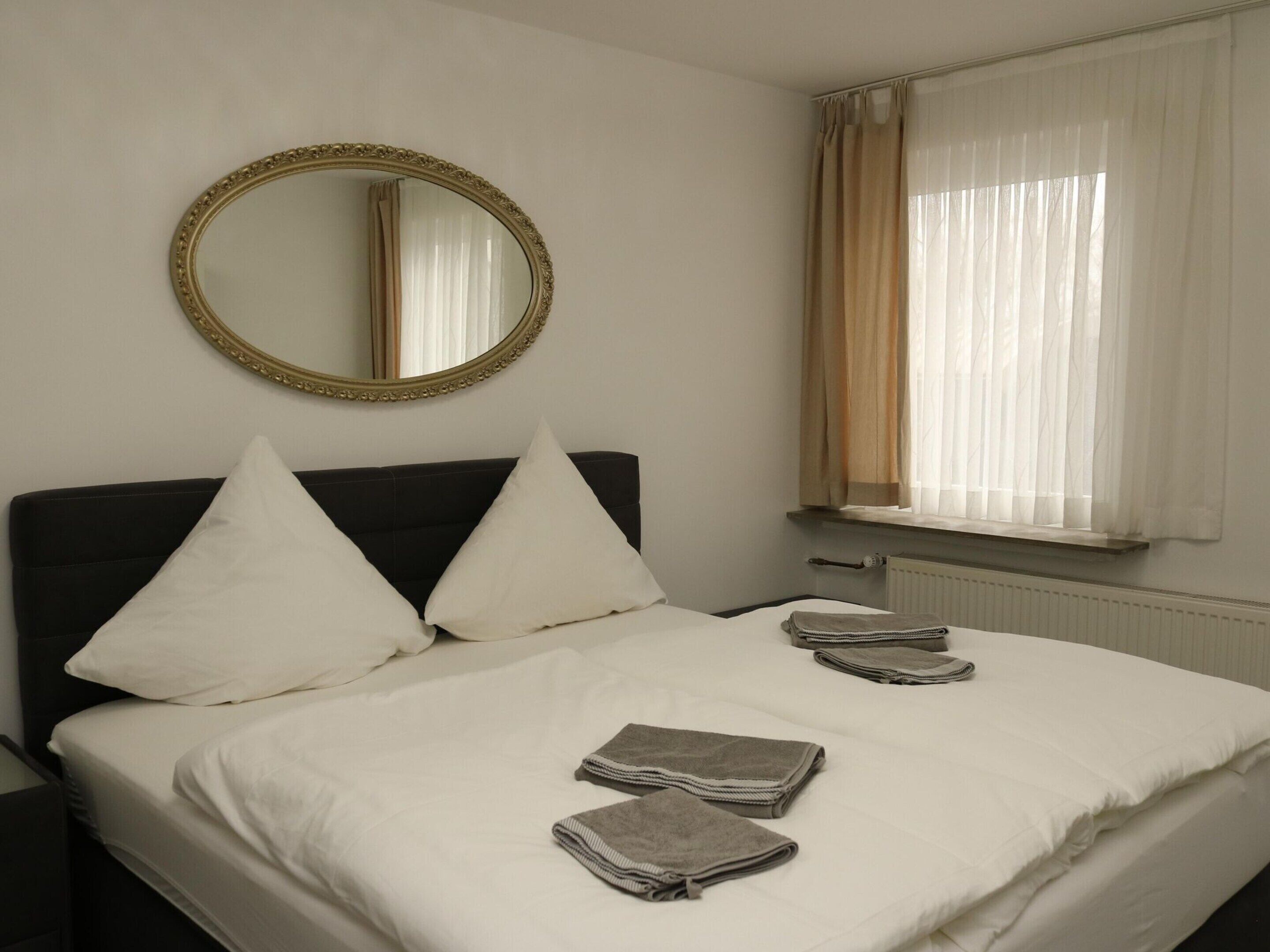 Quarto de hotel com cama, duas almofadas, um espelho redondo e uma janela com cortinas.