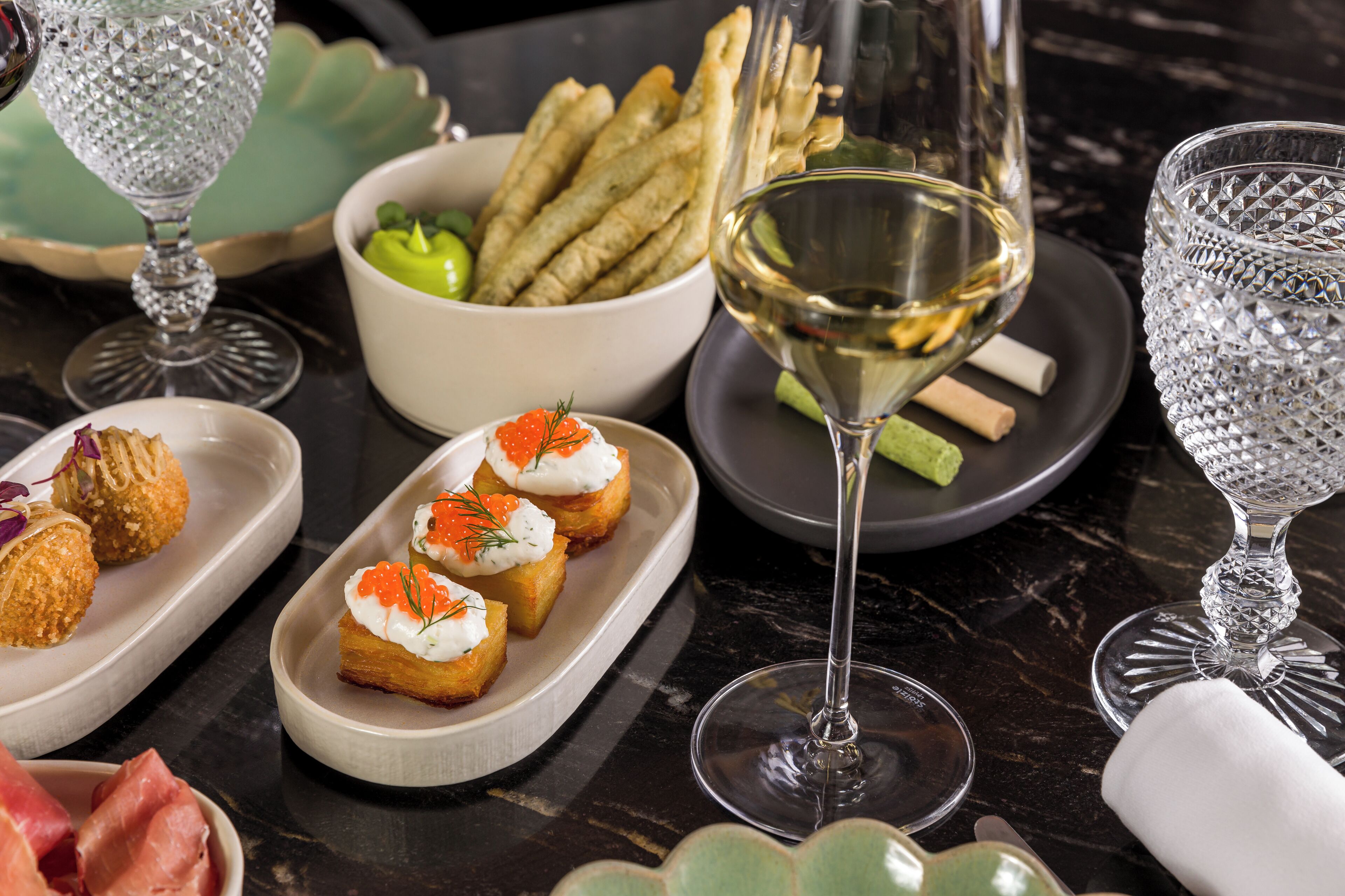 Uma mesa posta com diversos pratos, incluindo pão, caviar e uma taça de vinho branco.