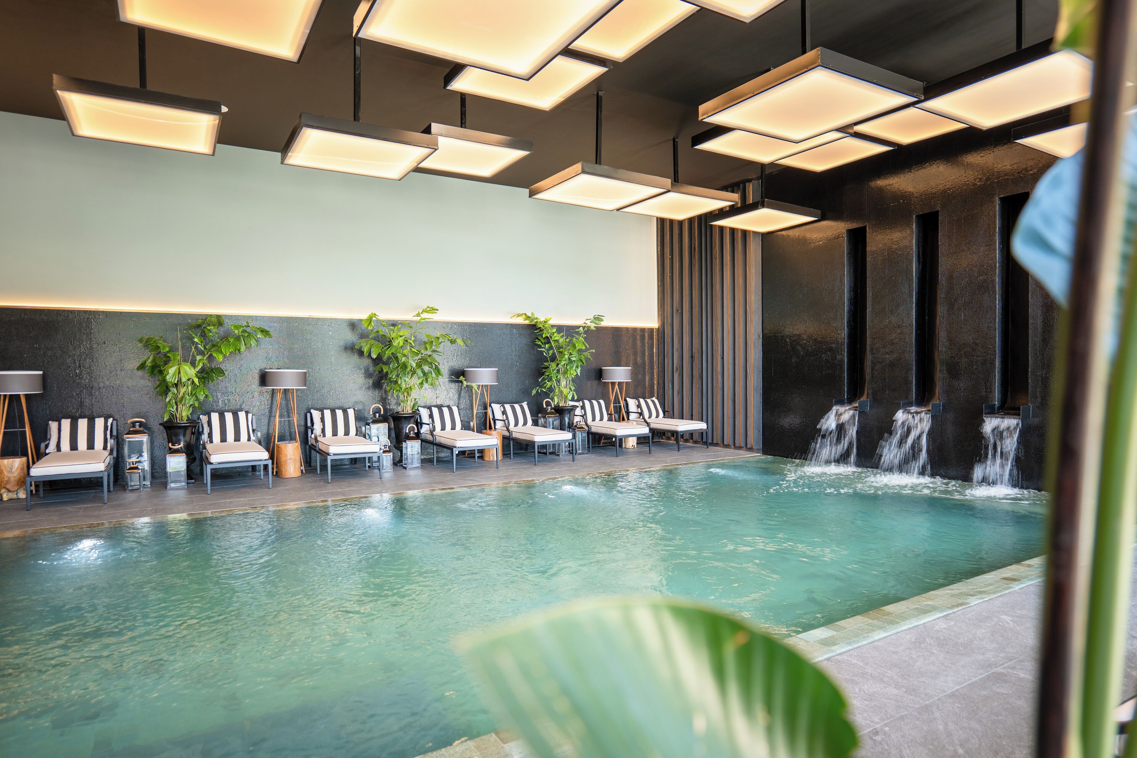 Uma área interna de piscina moderna com espreguiçadeiras, plantas em vasos e uma cascata.