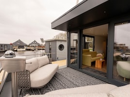 Um terraço moderno ao ar livre com um sofá e uma pequena mesa, uma porta de vidro que dá para um espaço interno com um sofá e uma mesa de centro, e uma vista de barcos e casas ao fundo.