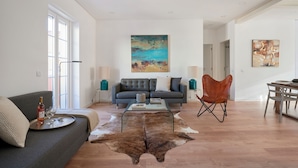 Uma sala de estar moderna com um sofá cinzento, uma mesa de centro e uma poltrona de couro. Há um quadro na parede e uma porta de vidro que dá para o exterior.