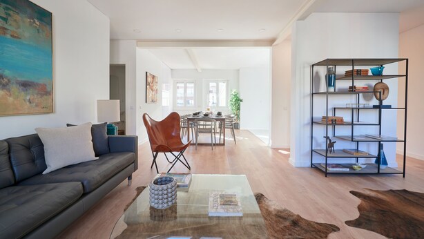 Uma sala de estar moderna com um sofá de couro, mesa de centro de vidro e um tapete de couro de vaca. Ao fundo, há uma área de jantar com mesa e cadeiras.