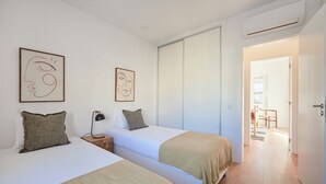 Quarto de hotel com duas camas, um guarda-roupa, uma escrivaninha com abajur e uma porta para um corredor.