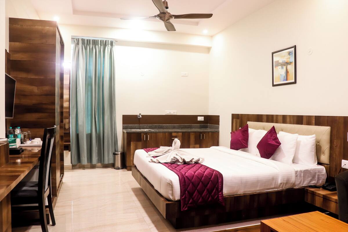 Quarto de hotel com cama, escrivaninha, cadeira, ventilador de teto, janela com cortinas e um quadro na parede.