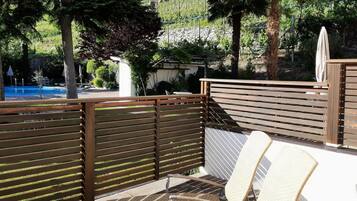 Uma área externa ensolarada com espaço para sentar, paredes de ripas de madeira, piscina e vegetação exuberante ao fundo.