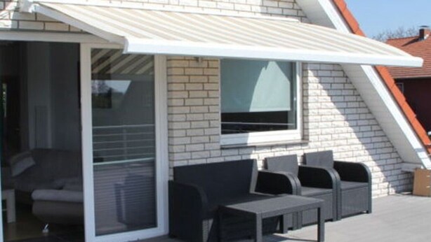 Uma casa moderna com telhado inclinado, um alpendre coberto com móveis de exterior e uma porta de correr que dá para uma varanda.