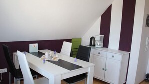 Uma sala de jantar com uma mesa branca, quatro cadeiras, um assento verde, um armário branco com uma cafeteira e uma pequena planta.