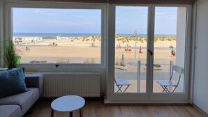 Quarto com uma janela grande com vista para uma praia, um sofá cinzento, uma mesa redonda pequena e duas cadeiras de metal.