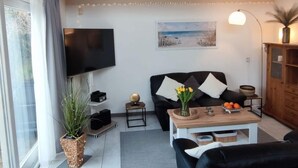 Uma sala de estar com uma TV de tela plana, um quadro de uma praia, um armário de madeira e uma mesa de centro com um vaso de flores.