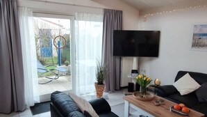 Sala moderna com sofá de couro preto, mesa de centro de madeira com um vaso de flores, TV de tela plana na parede e porta de vidro deslizante para um pátio.