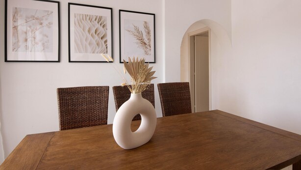 Uma mesa de jantar com um vaso branco e plantas secas, cercada por cadeiras de vime. Três obras de arte emolduradas estão penduradas na parede acima.