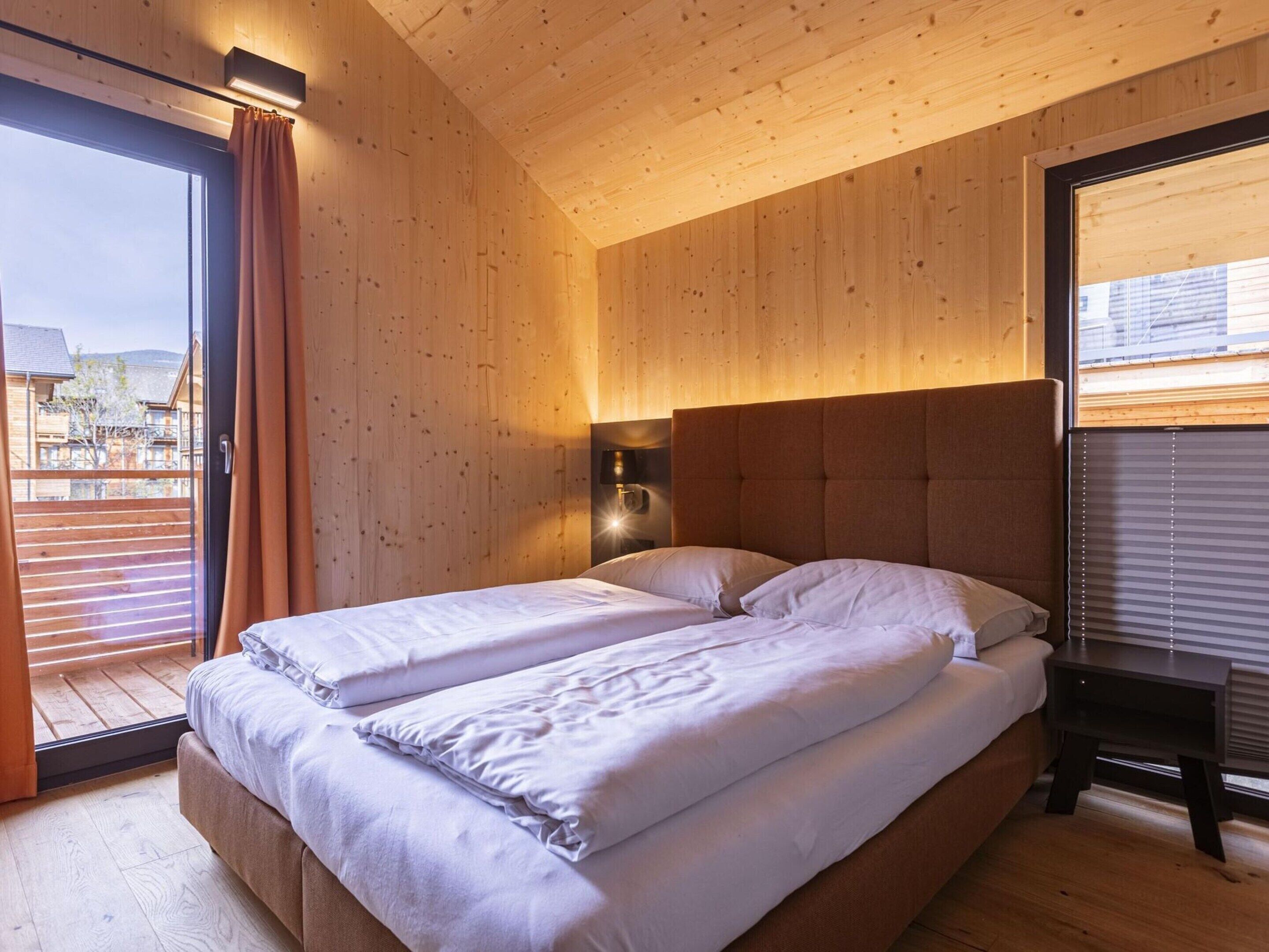 Quarto com teto de madeira, uma janela grande com cortinas, uma cama com roupa de cama branca e uma mesa de cabeceira.