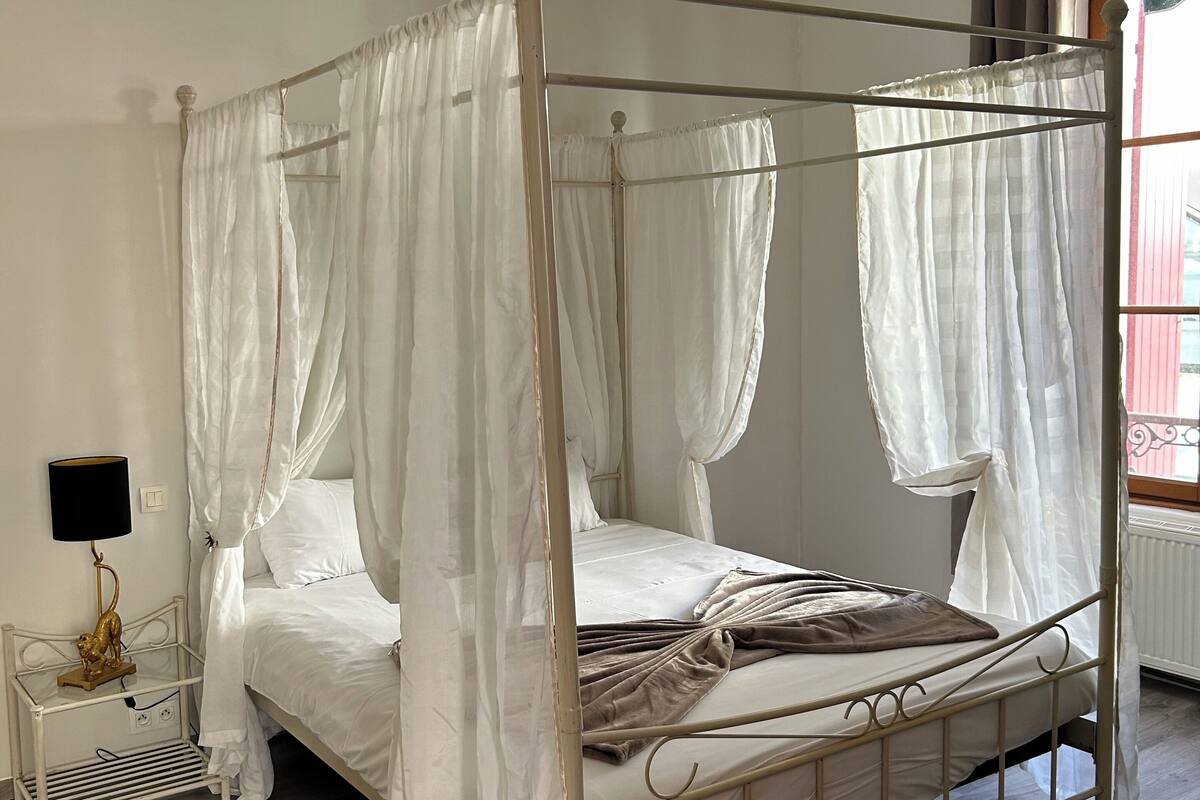 Uma cama de dossel com cortina, roupa de cama branca, mesa de cabeceira com abajur e janela com cortinas.