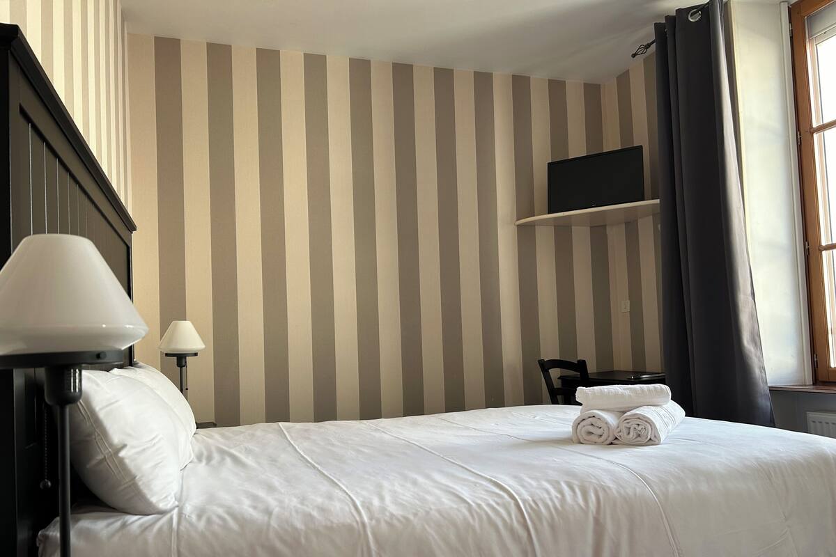Quarto de hotel com uma cama, uma televisão em uma estante, uma cadeira e uma janela com cortinas.
