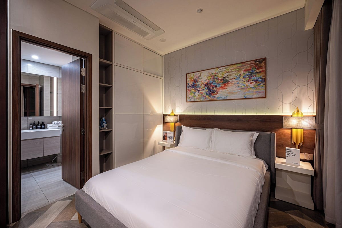 Quarto de hotel moderno com uma cama grande, mesa de cabeceira com luminária, um quadro na parede, e banheiro visível através de uma porta aberta.