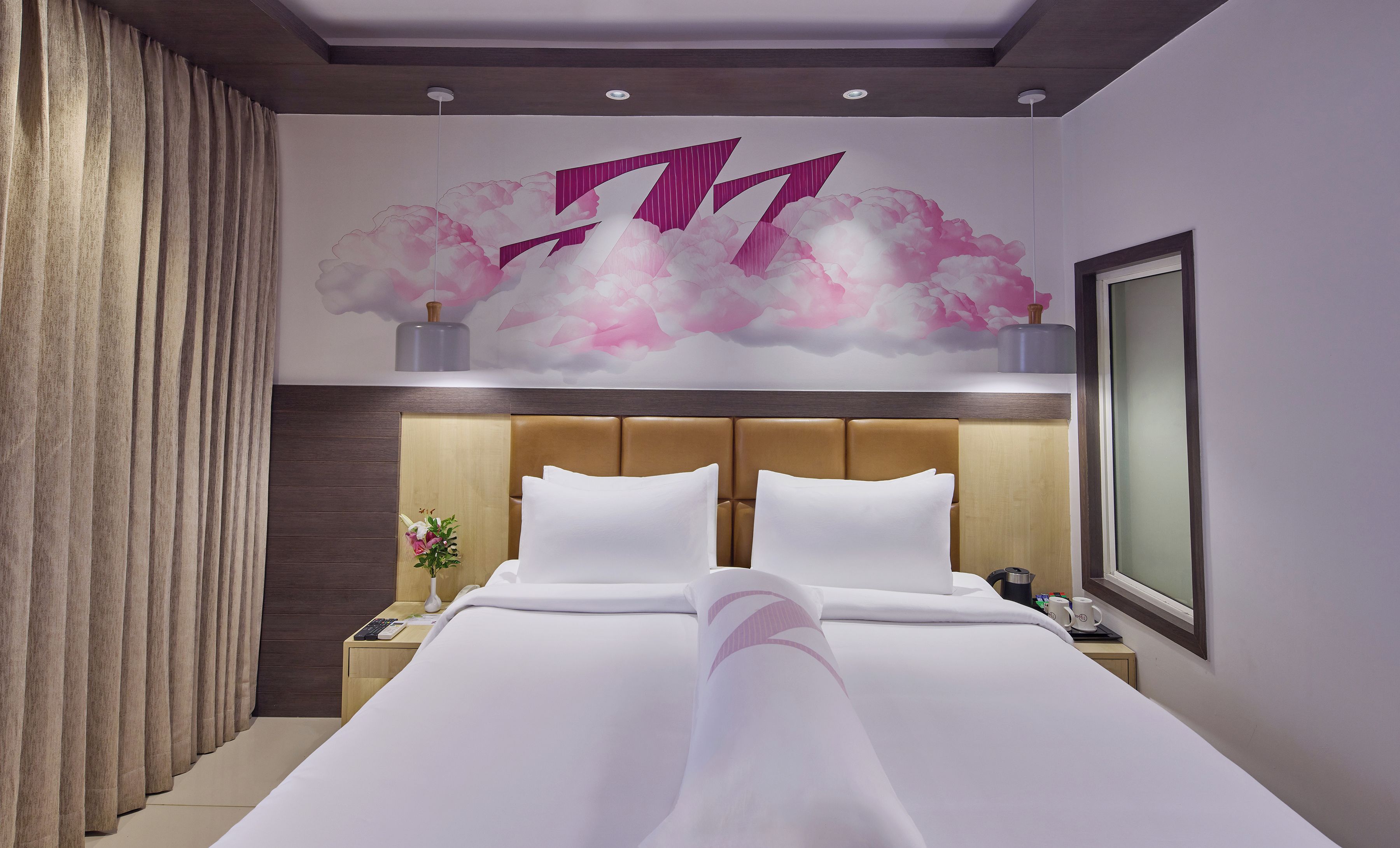 Quarto de hotel com uma cama grande, cabeceira com luminária e mesa de cabeceira com um vaso de flores, além de uma parede com um mural de nuvens.