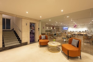 Um saguão de hotel com escada, área de estar com cadeiras laranja, mesa de centro e balcão de recepção com tela de televisão.