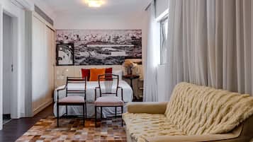 Quarto de hotel com piso xadrez, cama com mural de paisagem urbana, sofá e uma cadeira.