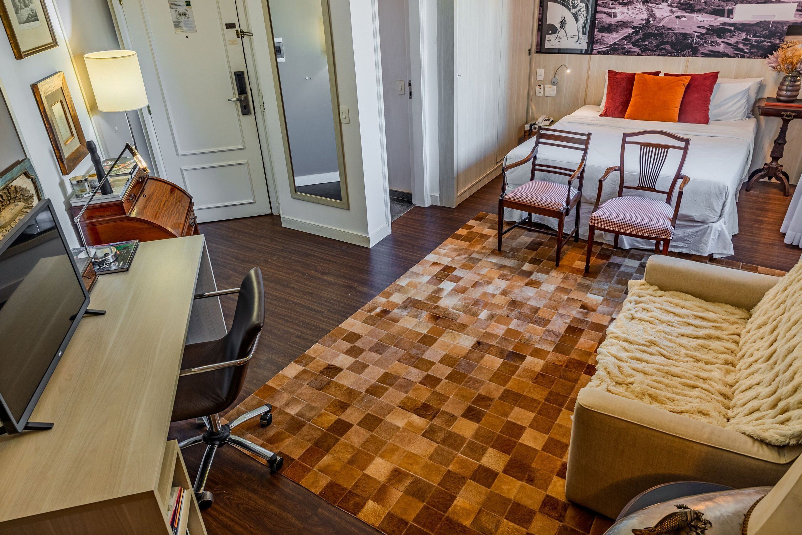 Quarto de hotel com uma mesa, cadeiras, cama, tapete xadrez e um quadro na parede.