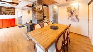 Uma sala de jantar com mesa e cadeiras de madeira, lareira de pedra e cozinha ao fundo.