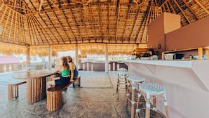 Uma área de estar externa com telhado de palha, móveis de madeira e um bar com banquetas.