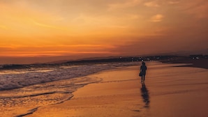 Cena de praia com uma pessoa caminhando pela costa ao pôr do sol.