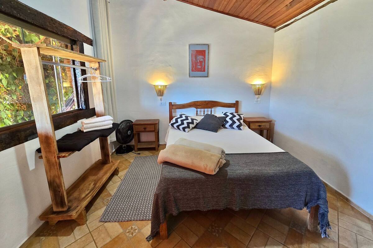 Quarto com cama de madeira, duas mesas de cabeceira com abajures, um banco e janela com cortinas.