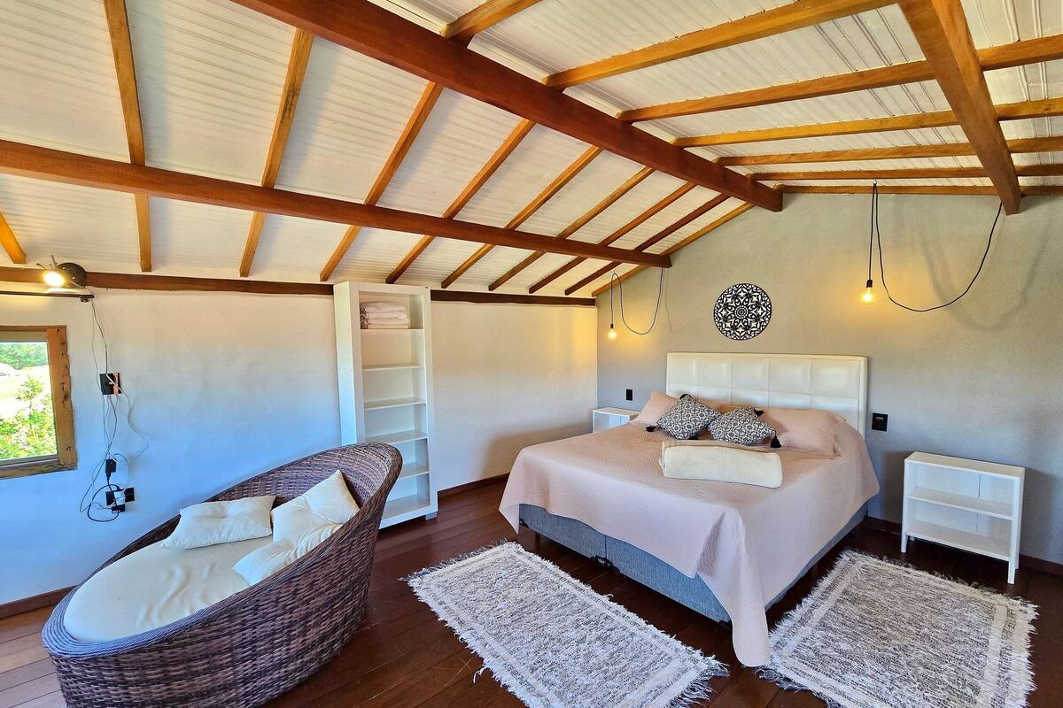 Quarto com cama, mesas de cabeceira, uma cadeira de vime, prateleira e janela.
