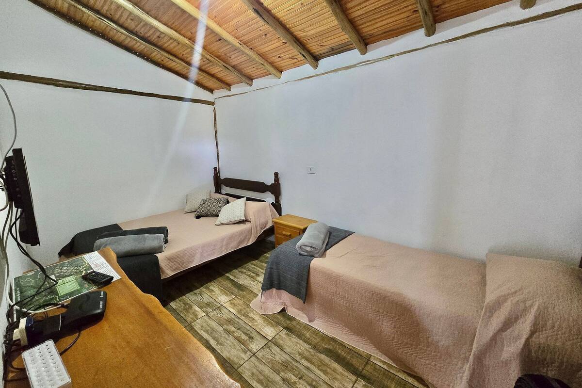 Quarto pequeno e simples, com duas camas individuais, uma escrivaninha de madeira e instalações elétricas básicas.