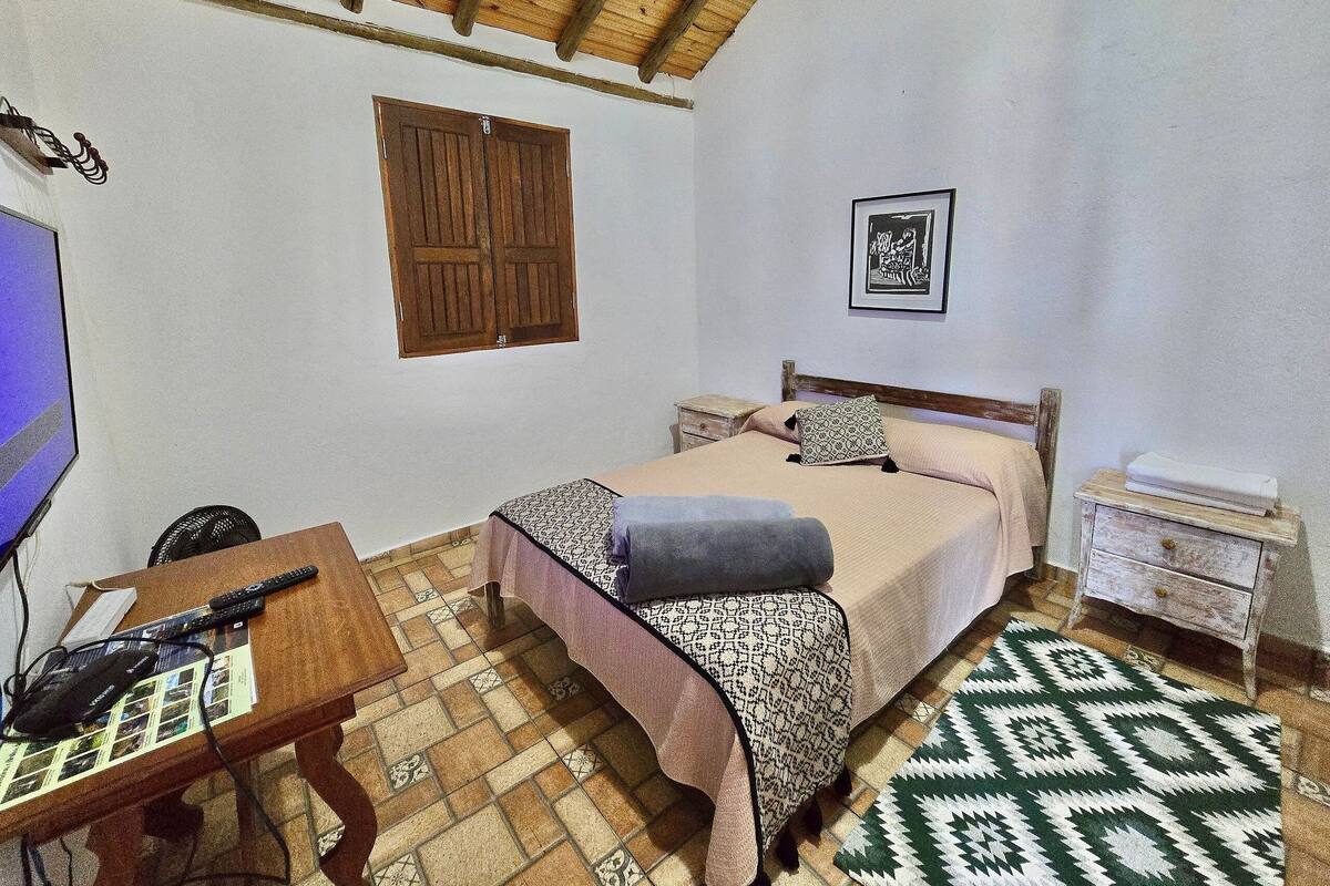 Quarto com cama, mesa de cabeceira de madeira, televisão e tapete estampado.