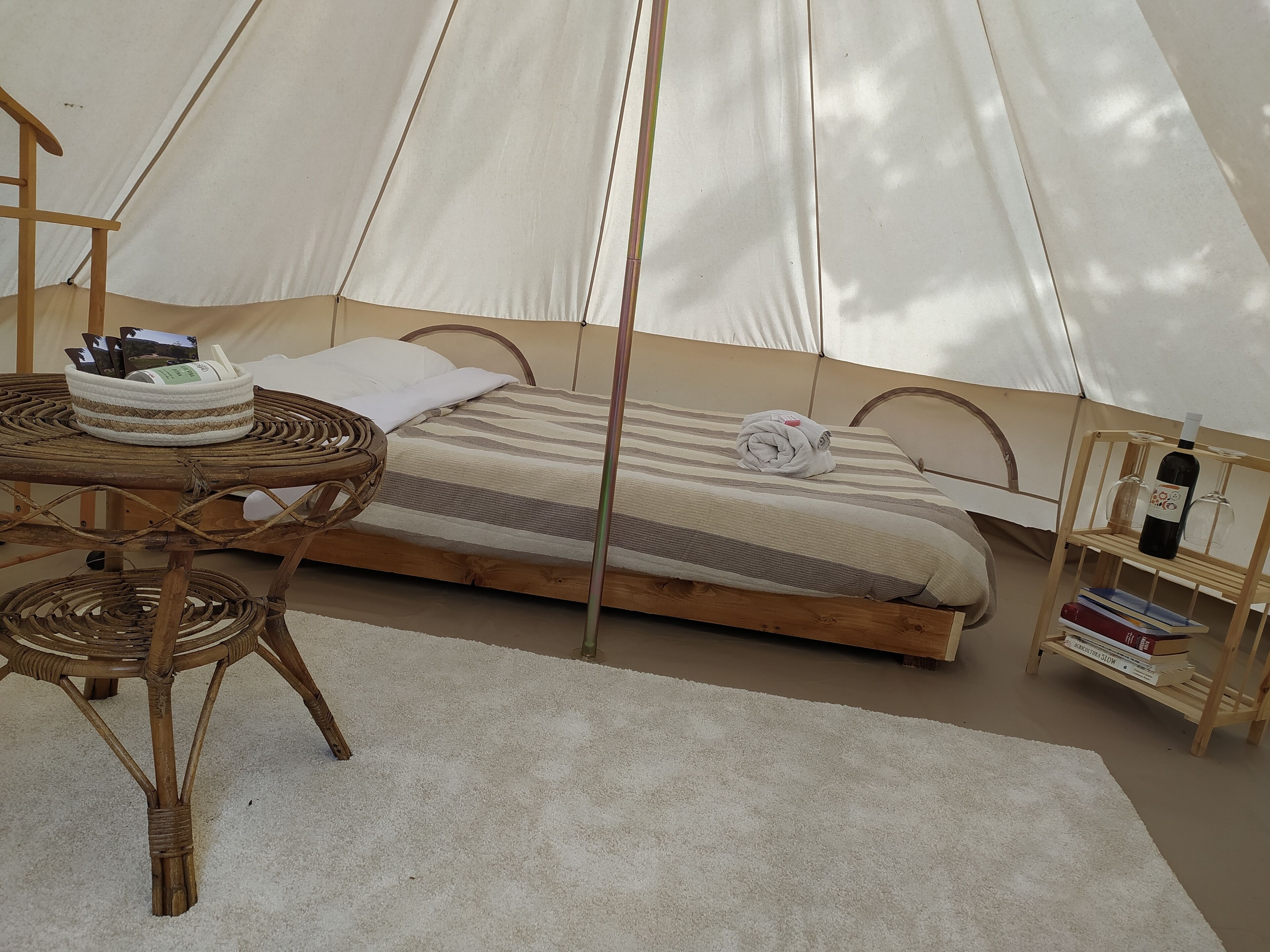 Um quarto em tenda aconchegante, com uma cama, uma mesa com revistas, uma garrafa de vinho e uma estante de livros.