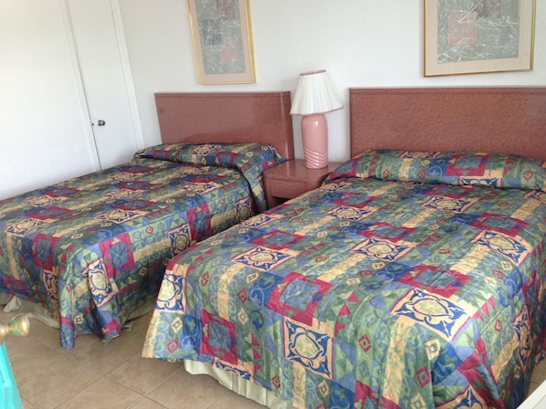 Quarto com duas camas de solteiro, uma mesa de cabeceira com abajur e uma porta.