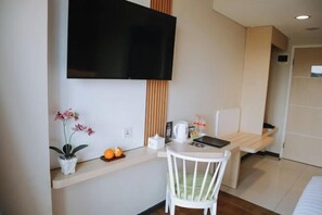 Quarto de hotel compacto com televisão de tela plana na parede, uma pequena mesa branca com uma cadeira, uma planta em vaso e uma cesta de frutas.