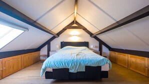 Quarto com teto inclinado, piso de madeira, cama com colcha azul clara e claraboia.