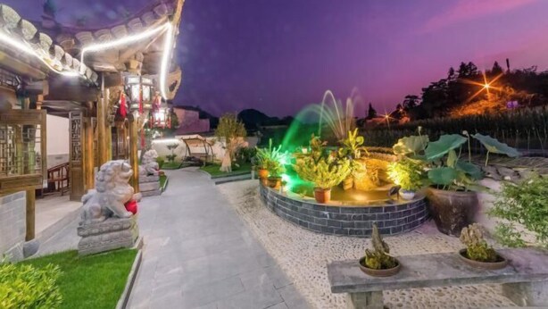 Uma área de jardim bem iluminada, com uma fonte, leões de pedra e uma estrutura arquitetônica tradicional.