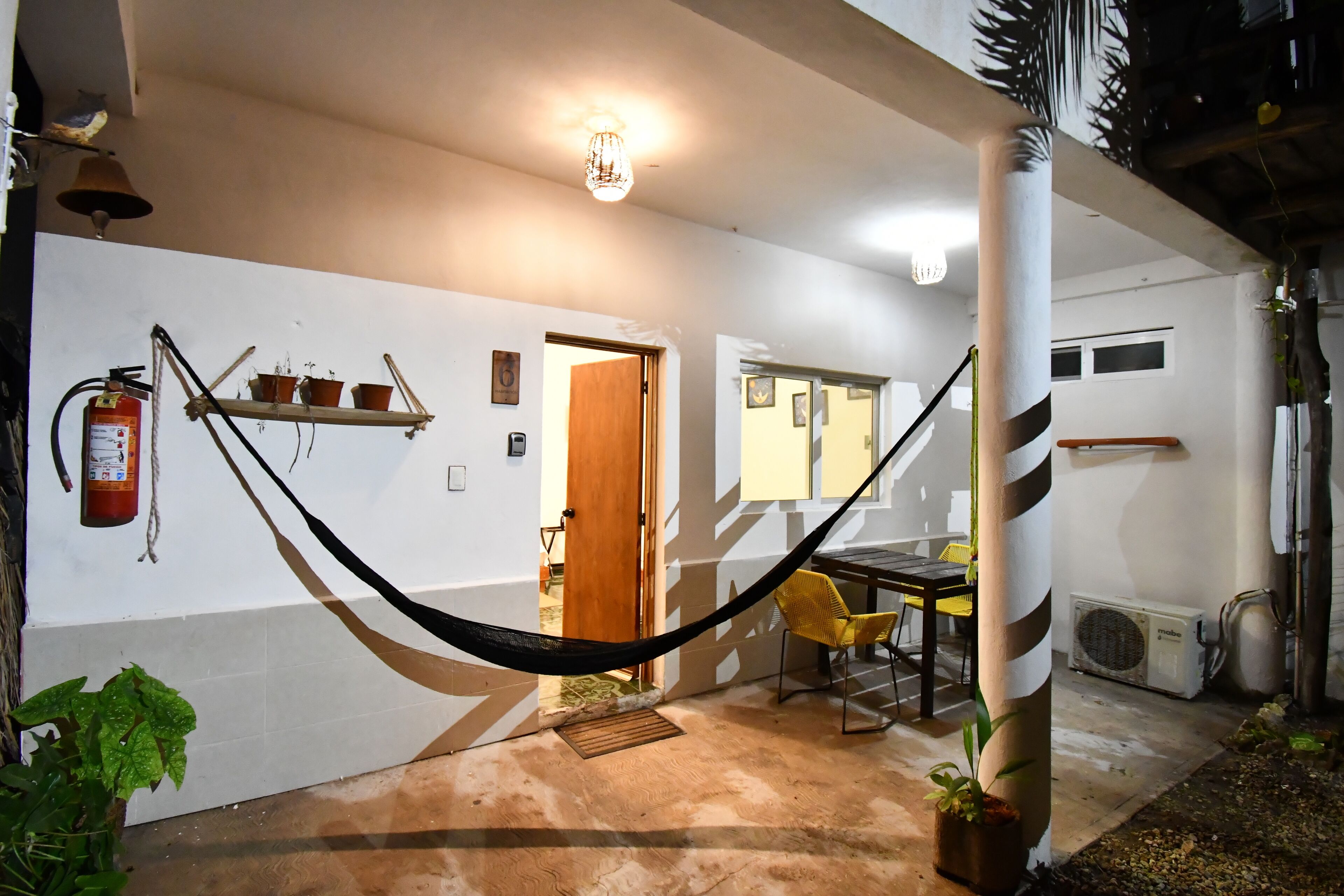 Quarto com rede, extintor de incêndio, prateleira com plantas em vasos, cadeira amarela e mesa.