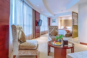 Quarto de hotel com uma cama grande, duas poltronas, uma mesa de centro com frutas e uma televisão em um suporte.