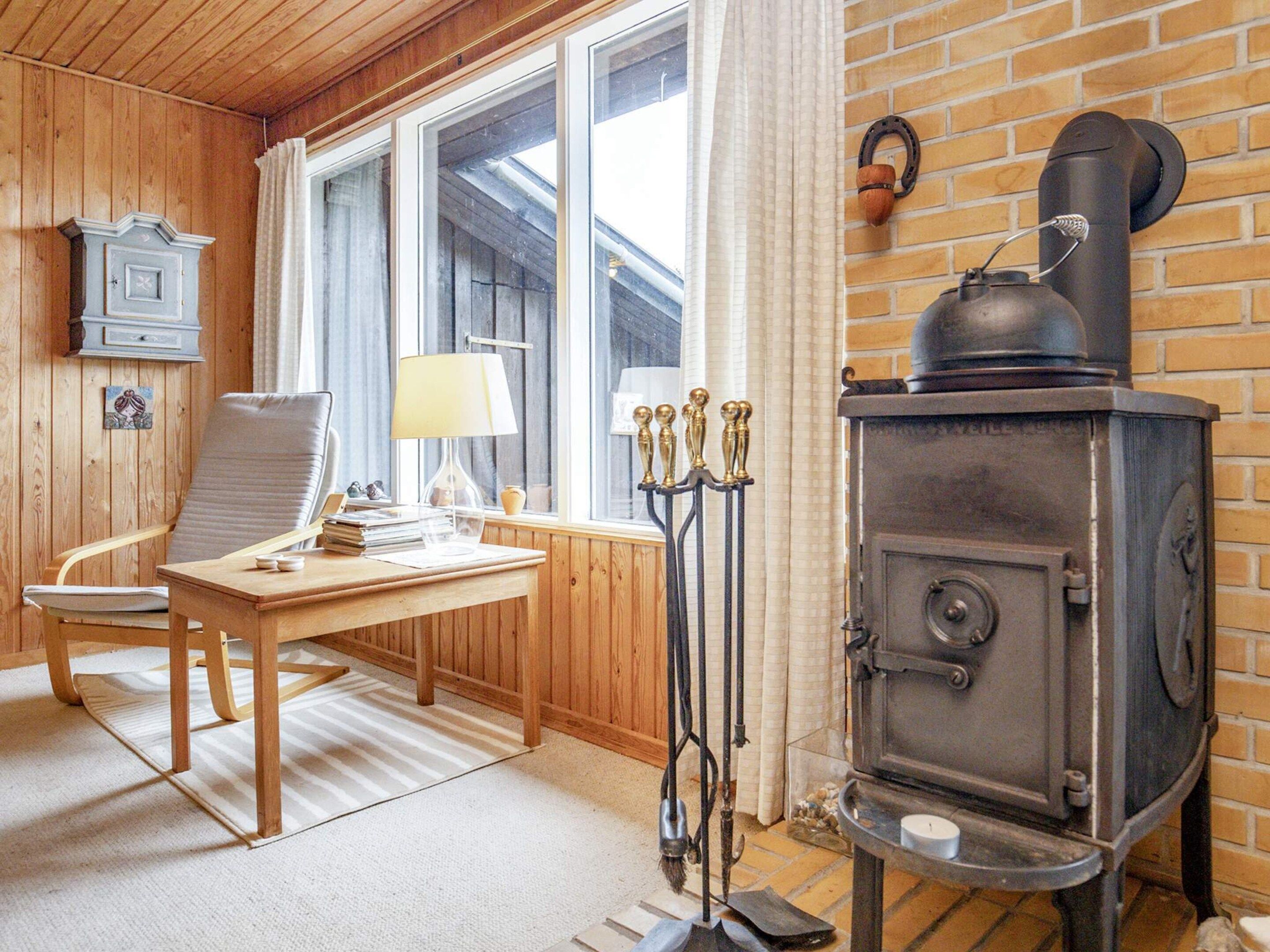 Um quarto aconchegante com decoração em madeira, uma janela grande, uma mesa com luminária, uma cadeira e um fogão antigo.