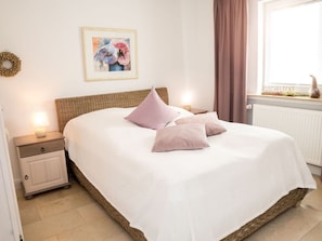 Uma cama bem arrumada com almofadas brancas e lavanda, um cabeceiro de vime, uma mesa de cabeceira com abajur e uma obra de arte floral emoldurada na parede.